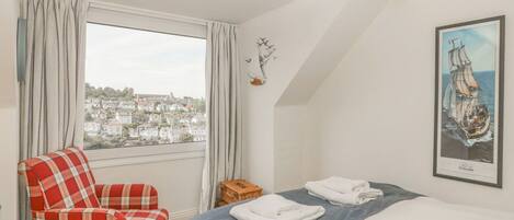 Um quarto com uma cama, uma poltrona xadrez vermelha, uma janela com vista para a cidade, um quadro emoldurado de um navio e uma pequena mesa de madeira com uma cesta.