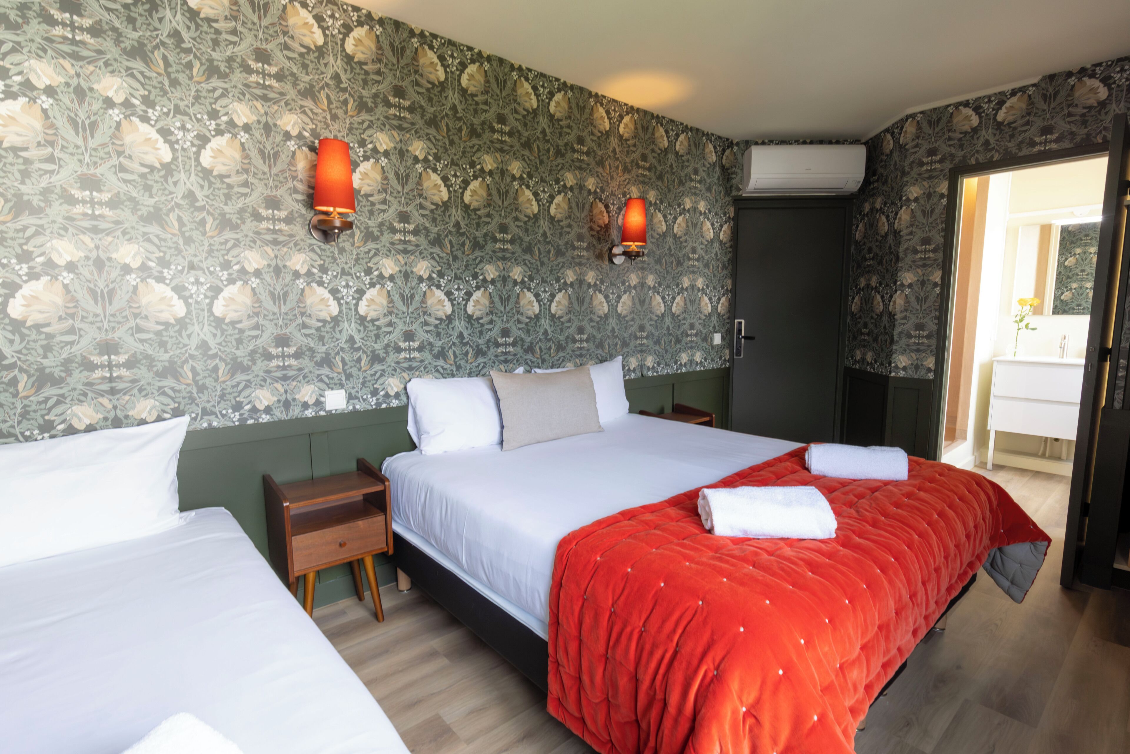 Quarto de hotel com uma cama grande, uma mesa de cabeceira, uma luminária na parede e uma porta para o banheiro.