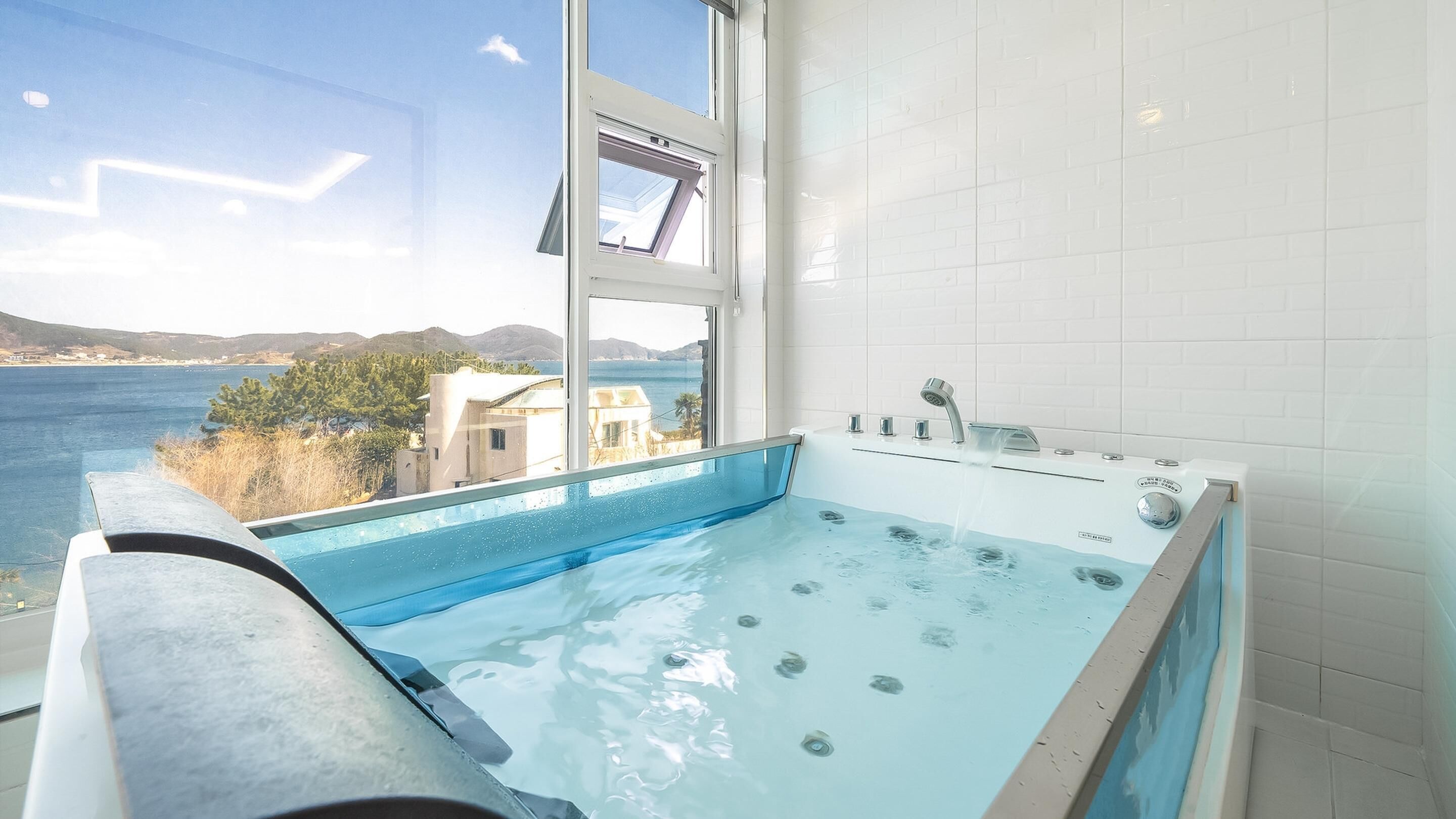 Um banheiro moderno com uma banheira grande, uma janela com vista para o mar e colinas, e paredes com azulejos brancos.