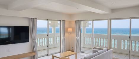 Uma sala de estar moderna com uma TV de tela plana, uma mesa de centro de madeira, um sofá cinza e uma luminária. Grandes janelas oferecem vista para o mar.