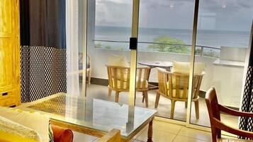 Quarto de hotel com cama, móveis de madeira, uma mesa de vidro e vista para o mar através de grandes janelas.