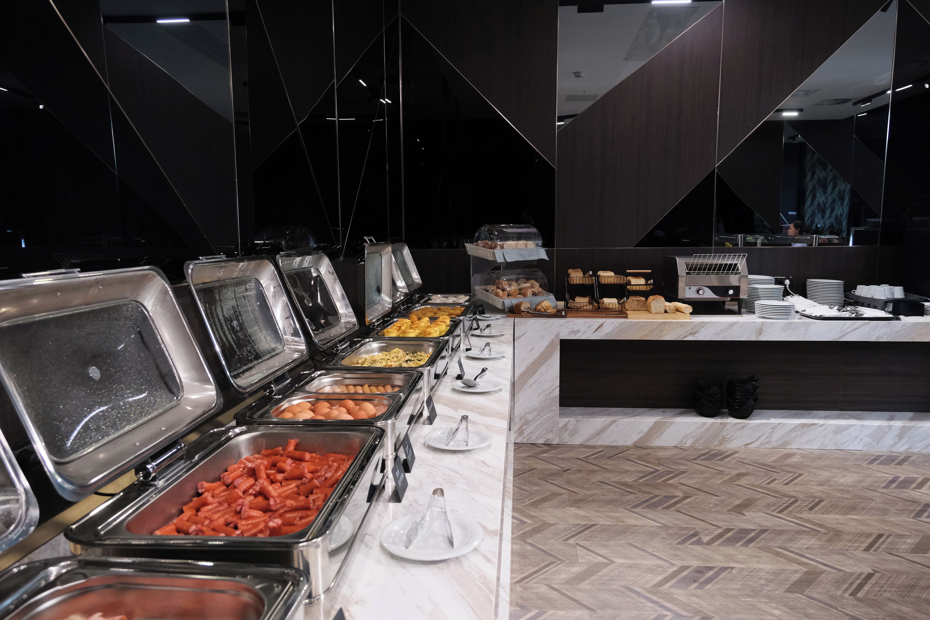 Disposição de buffet com diversos alimentos em bandejas, balcão com pratos e talheres, e design de interiores moderno.
