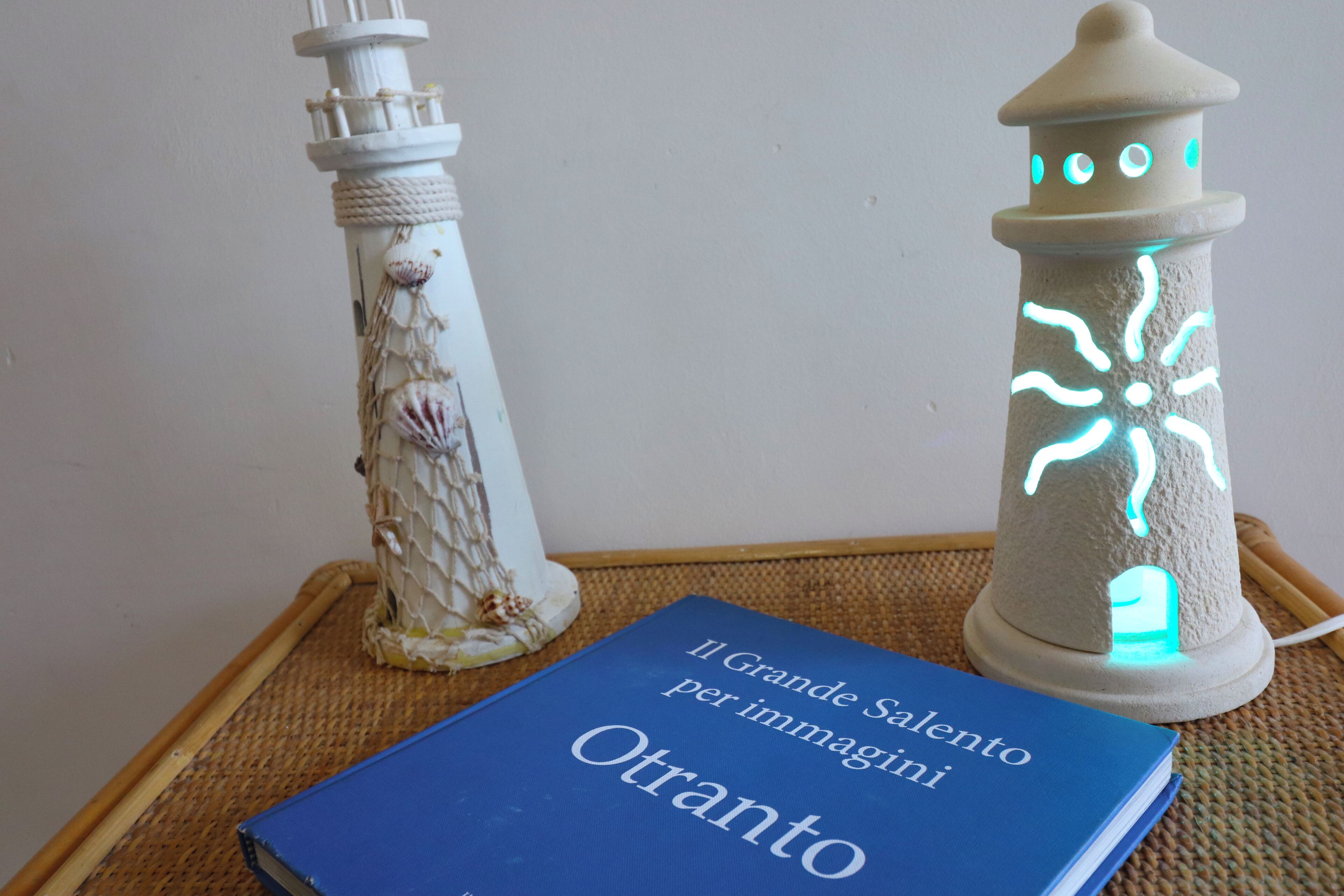 Um farol decorativo e um livro intitulado "Il Grande Salento per immagini" sobre uma superfície trançada.