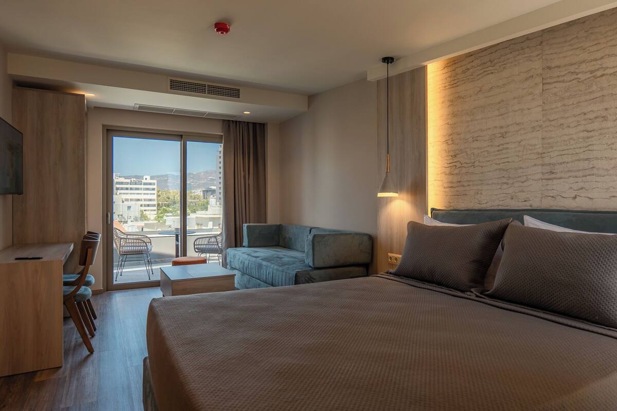 Quarto de hotel com uma cama grande, um sofá, uma escrivaninha e uma varanda com vista para a cidade.