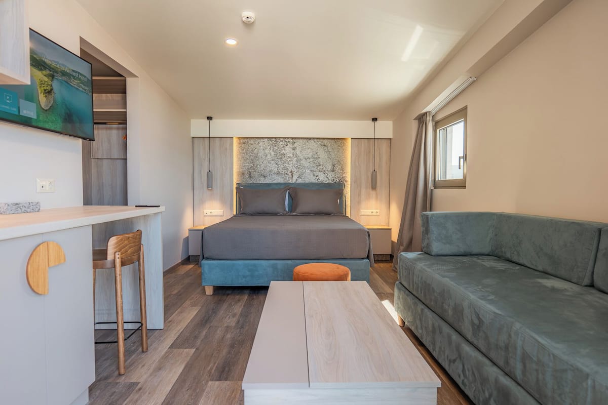 Quarto moderno e compacto com cama, sofá, mesinha e televisão fixada na parede.