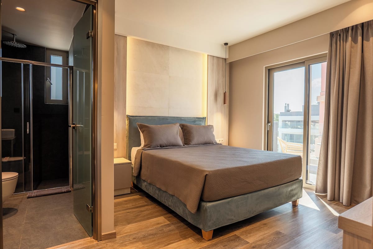 Quarto moderno com cama grande, chuveiro em vidro e vista para a cidade.