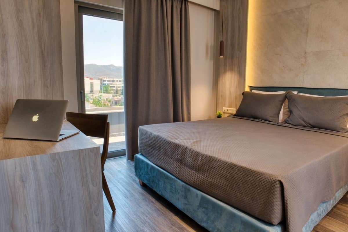 Quarto de hotel com cama, escrivaninha com laptop, vista da cidade e uma janela grande.