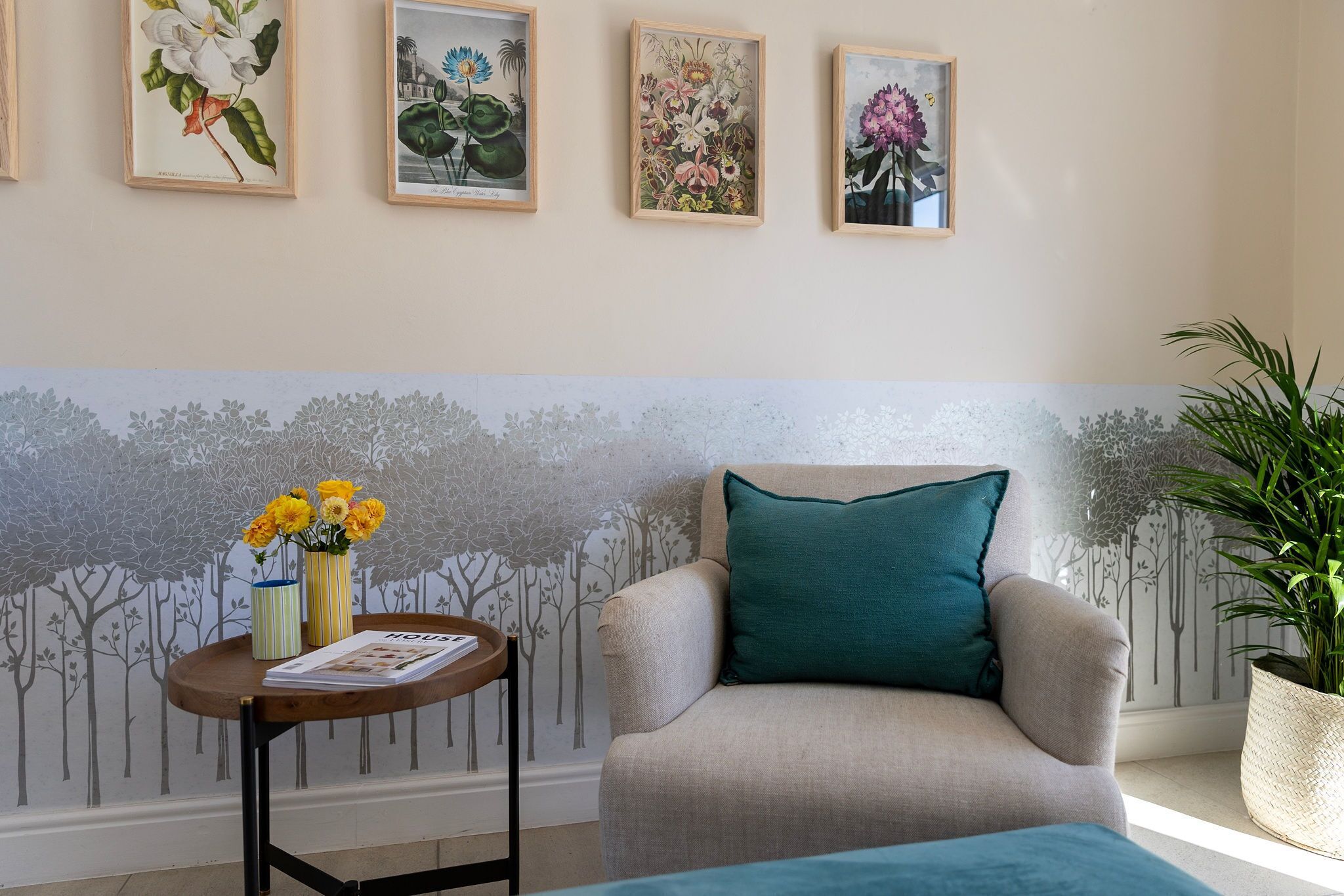 Um quarto com um sofá bege, uma mesa redonda de madeira com um vaso de flores amarelas e uma parede com um mural de uma árvore.