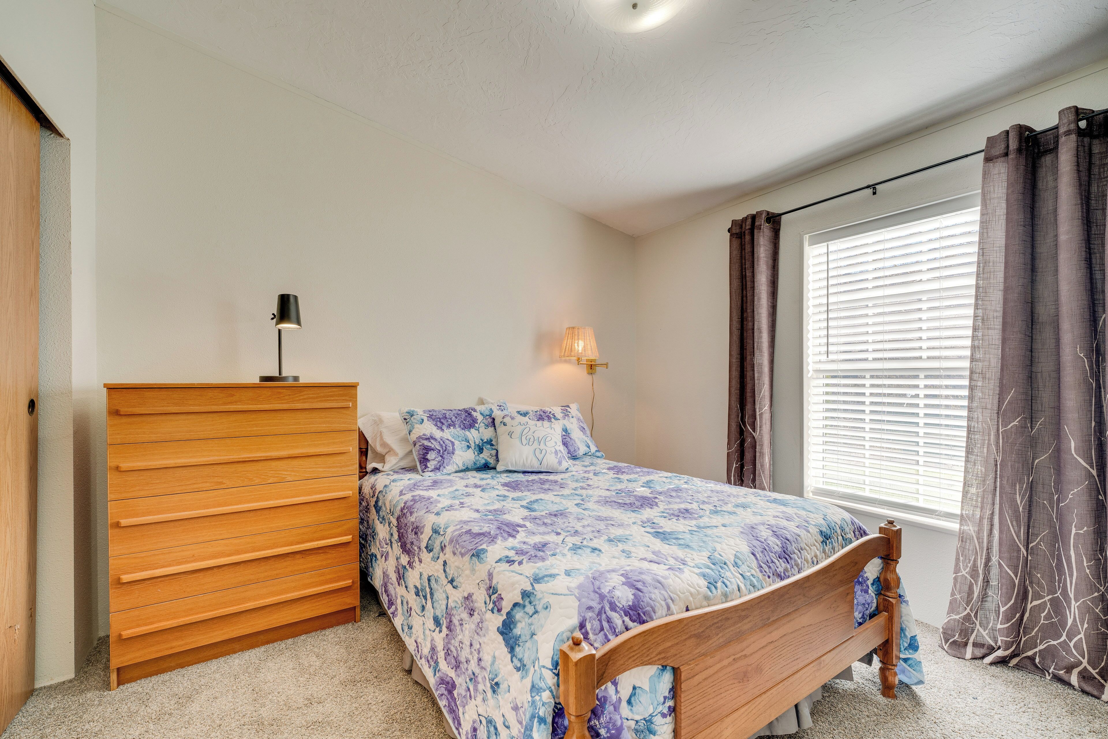 Quarto com cômoda de madeira, cama com colcha florida, luminária na parede e janela com persianas.