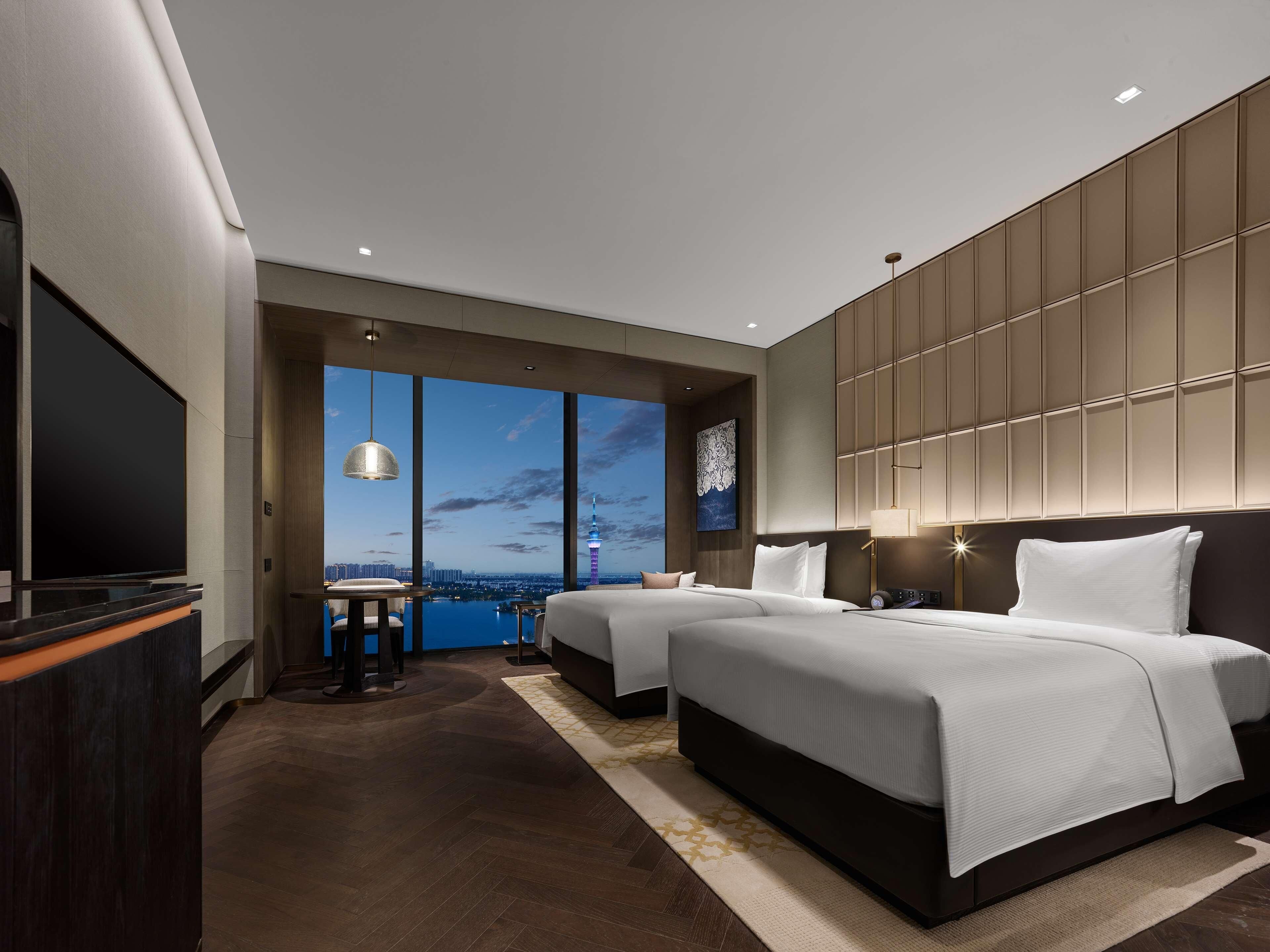 Quarto de hotel moderno com uma cama grande, TV e vista para o horizonte urbano por grandes janelas.