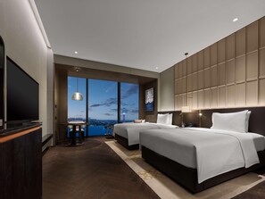 Quarto de hotel moderno com uma cama grande, TV e vista para o horizonte urbano por grandes janelas.
