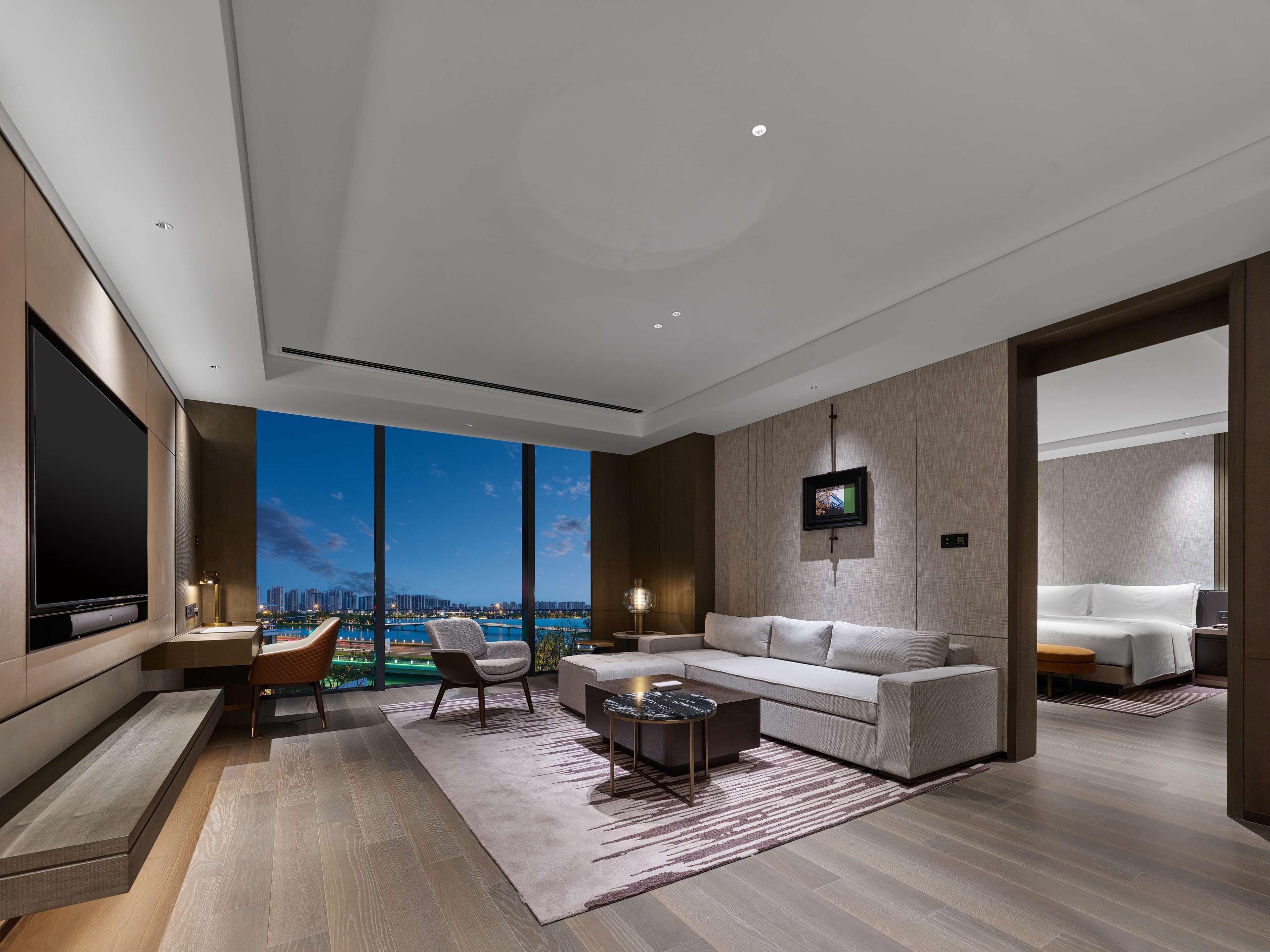 Uma sala de estar moderna com uma TV grande, assentos confortáveis, uma mesa de centro e vista para o horizonte da cidade ao entardecer.
