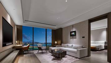 Uma sala de estar moderna com uma TV grande, assentos confortáveis, uma mesa de centro e vista para o horizonte da cidade ao entardecer.