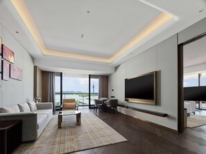 Uma sala de estar moderna com uma grande televisão de tela plana, um sofá, uma mesa de centro e vista para um horizonte urbano através de amplas janelas.