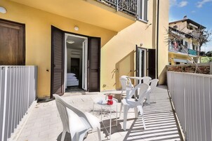 Um pátio ao ar livre banhado pelo sol, com cadeiras de plástico branco e uma mesa pequena, um edifício amarelo com persianas abertas e uma varanda com guarda-corpo.