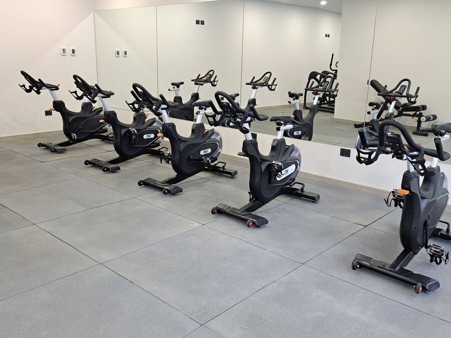 Uma academia com bicicletas ergométricas dispostas em linha, um espelho na parede e piso de azulejos.