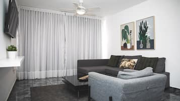 Uma sala de estar moderna com um sofá cinzento, uma mesa de centro de vidro e um ventilador de teto. Há duas gravuras botânicas emolduradas na parede.