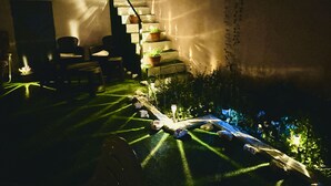 Escadaria com pouca luz, plantas em vasos, um banco de madeira e uma pequena mesa com uma vela.