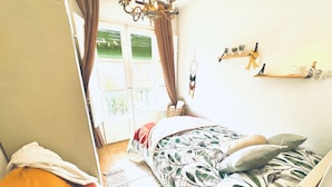 Quarto com cama, uma cadeira, prateleira com plantas e janela com cortinas.