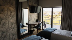 Quarto de hotel com duas camas, uma escrivaninha, uma cadeira, uma televisão e uma varanda com vista para a cidade.