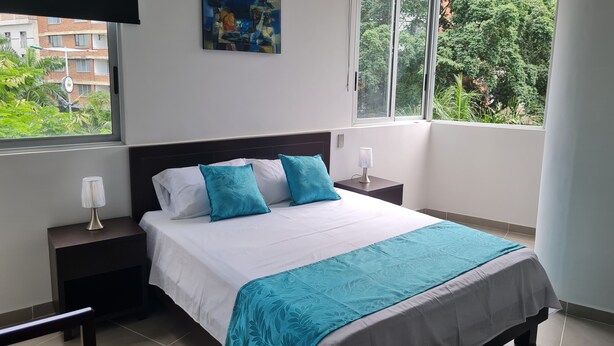 Quarto moderno com uma cama grande, duas mesas de cabeceira, um quadro e vista para árvores e edifícios pela janela.