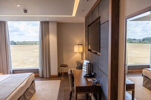 Quarto de hotel com uma cama grande, uma escrivaninha com abajur, uma TV na parede e vista para um campo pela janela.