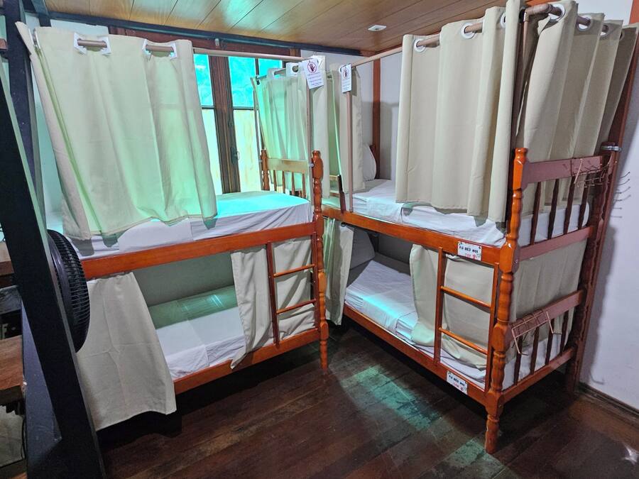 Quarto com beliches de madeira, cada um com colchão e travesseiro. Há uma escada entre as camas para acesso.