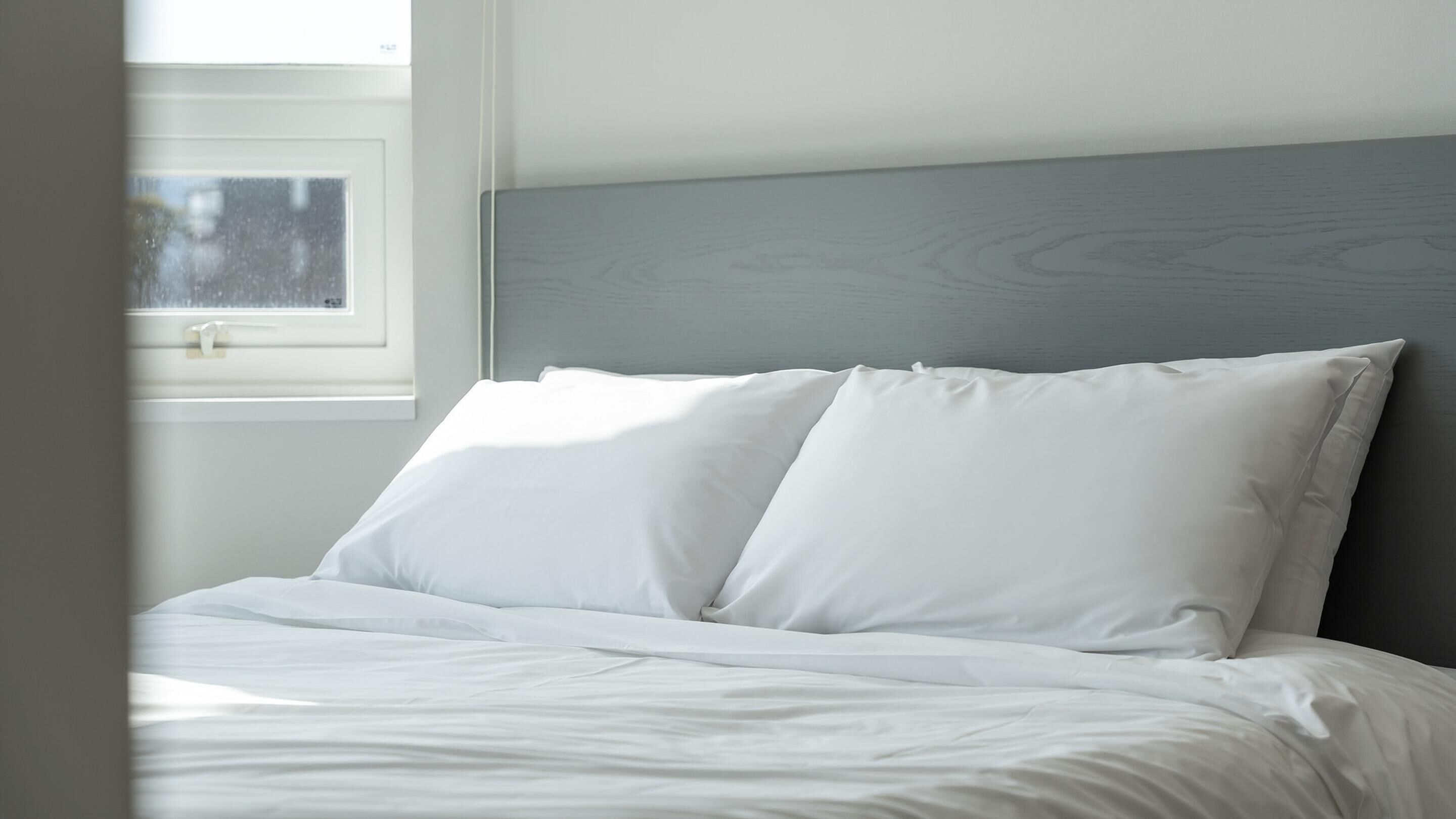 Uma cama bem arrumada com roupa de cama e almofadas brancas, cabeceira cinza e uma janela com persianas.