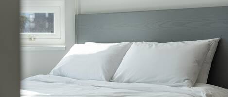 Uma cama bem arrumada com roupa de cama e almofadas brancas, cabeceira cinza e uma janela com persianas.
