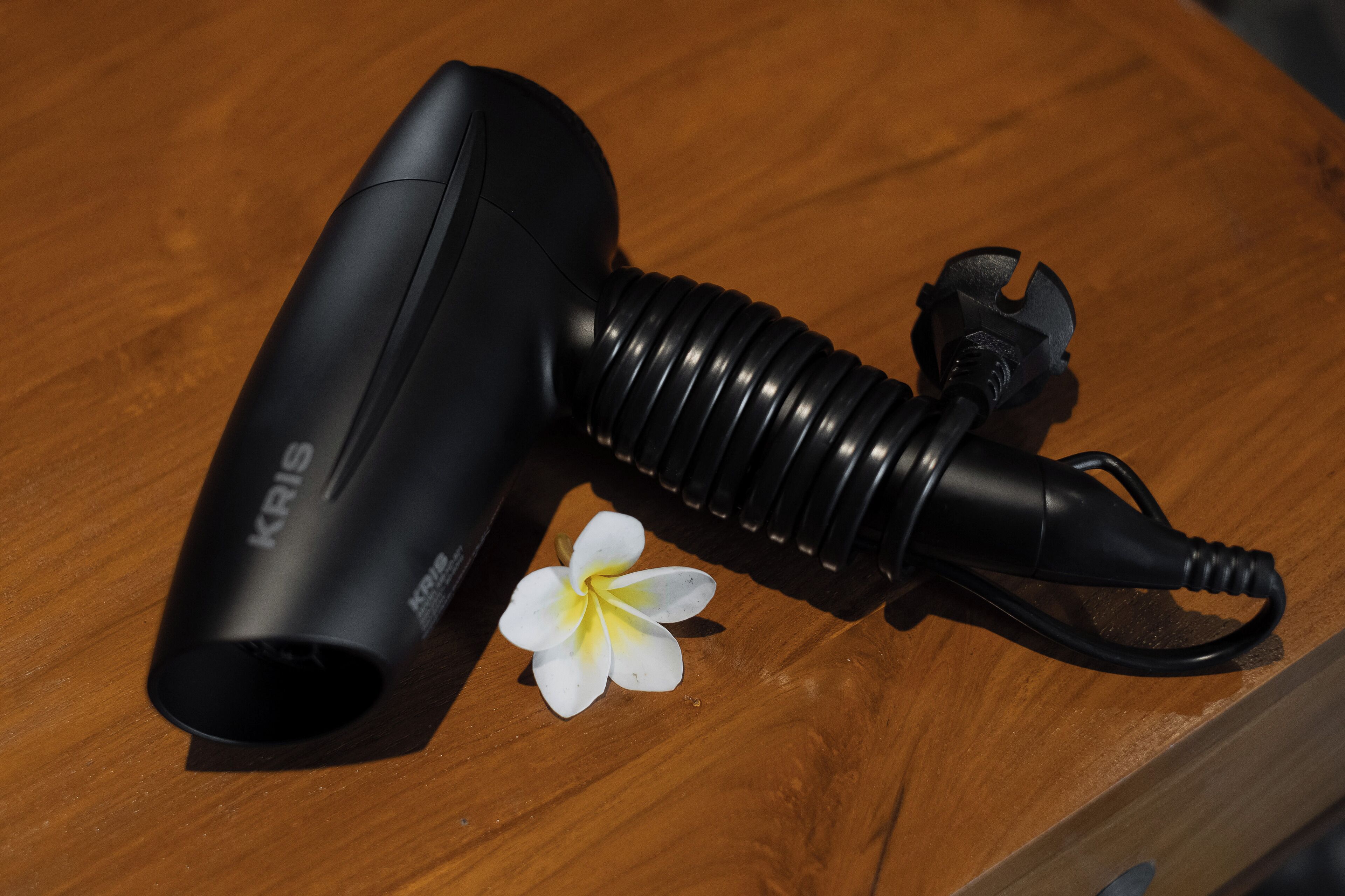 Secador de cabelo preto com tomada e uma flor branca sobre superfície de madeira.