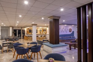Um saguão de hotel moderno com balcão de recepção, área de estar e um bar/lounge ao fundo.