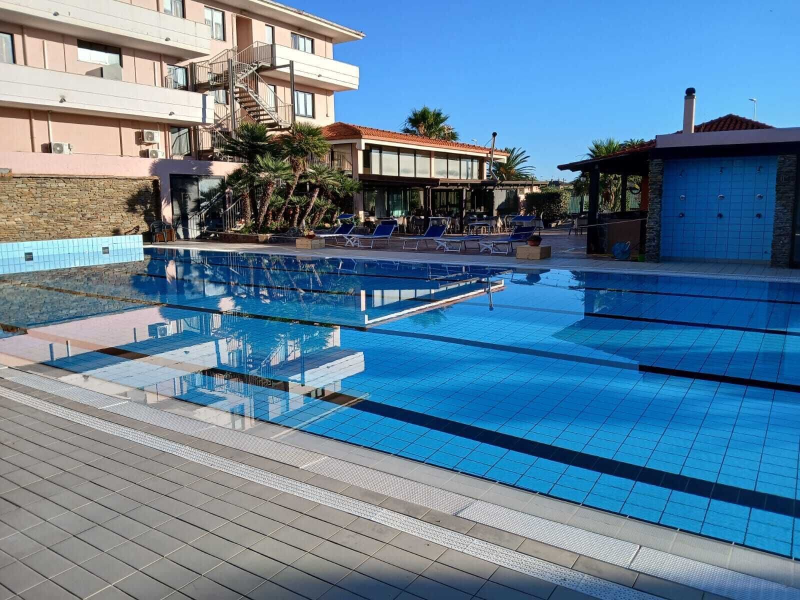 Piscina com água azul cristalina, rodeada por espreguiçadeiras e um edifício com varandas.