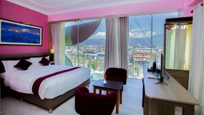 Quarto de hotel com cama, escrivaninha, cadeira e uma grande janela com vista para a cidade.