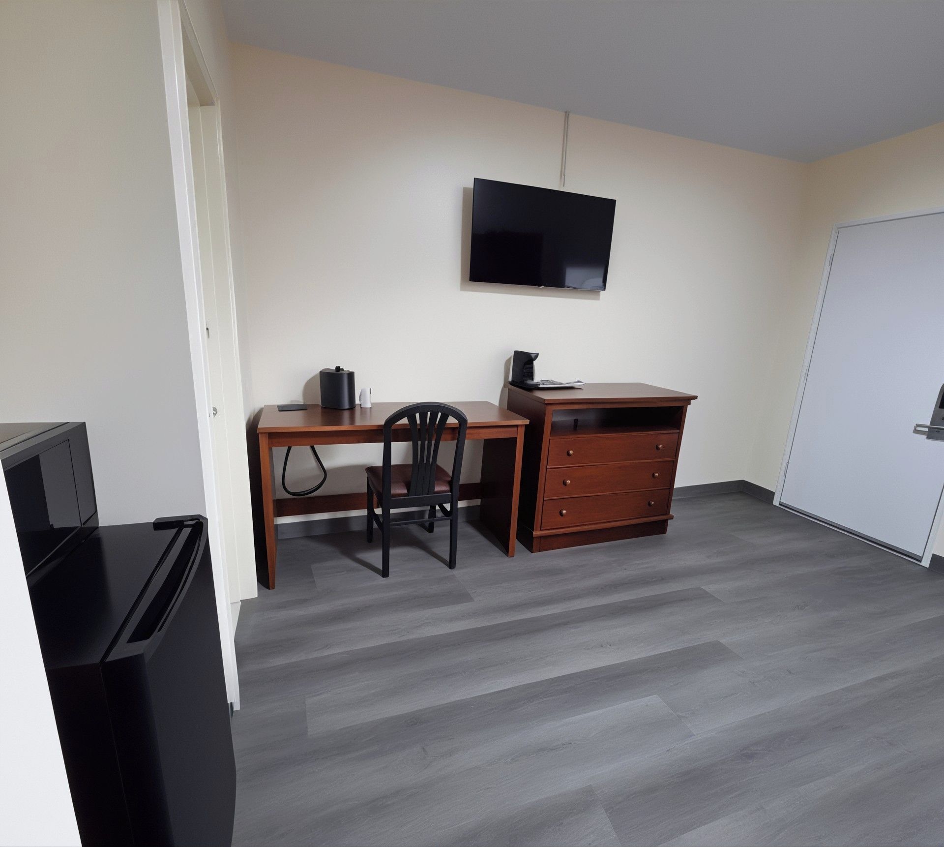Um quarto pequeno com escrivaninha, cadeira, televisão fixada na parede, armário e porta branca.