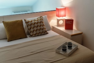 Cama bem arrumada com duas almofadas, mesa de cabeceira com abajur e um livro, e bandeja com duas xícaras.