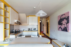 Uma cozinha compacta com armários brancos, micro-ondas e fogão. Há uma área de refeições com mesa e cadeiras brancas.