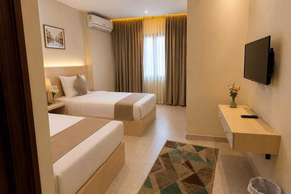Quarto de hotel com duas camas, televisão, escrivaninha, lixeira e janela com cortinas.