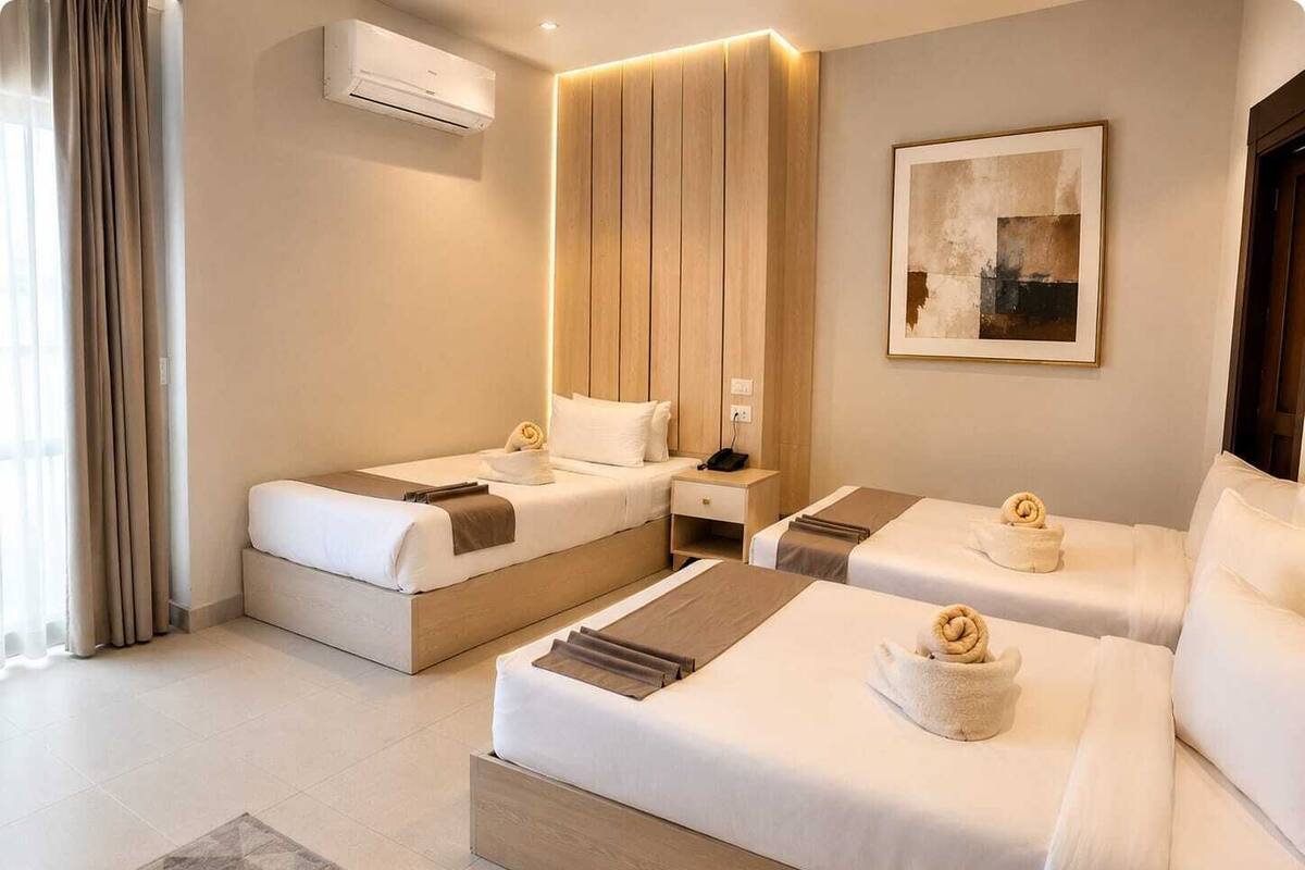Quarto de hotel com duas camas, uma mesa de cabeceira, ar condicionado, um quadro na parede e um tapete com padrão geométrico.