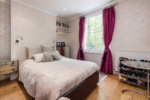 Quarto com cama, radiador, sapateira e janela com cortinas.