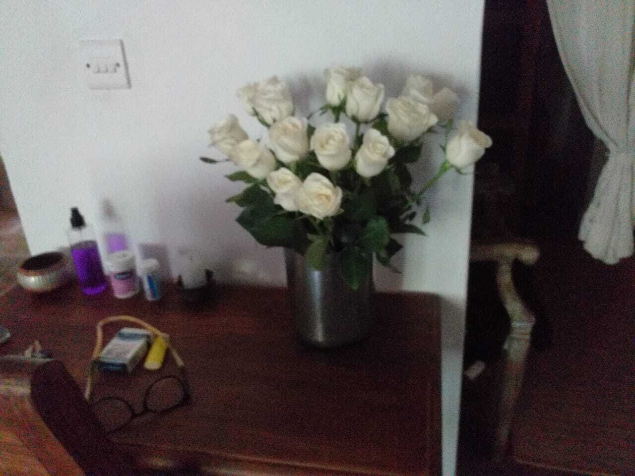 Uma pequena mesa com um vaso de metal contendo rosas brancas, um par de óculos e alguns objetos pequenos no lado esquerdo.