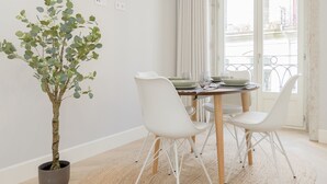Uma área de refeições com uma mesa redonda, cadeiras brancas, um tapete tecido e um vaso com planta.