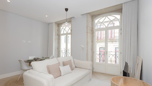 Uma sala de estar moderna com sofá branco, mesa de jantar e cadeiras. Há uma janela grande com vista para uma varanda.