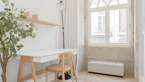 Quarto com escrivaninha branca, cadeira, planta em vaso, banco e janela com vista para o exterior.