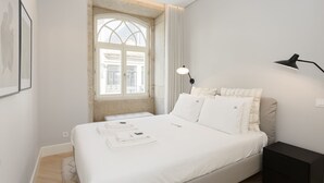 Um quarto com cama, mesa de cabeceira, abajur, janela e um quadro na parede.