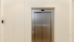 Um elevador moderno com acabamento metálico, situado em um corredor com paredes claras e chão de madeira.