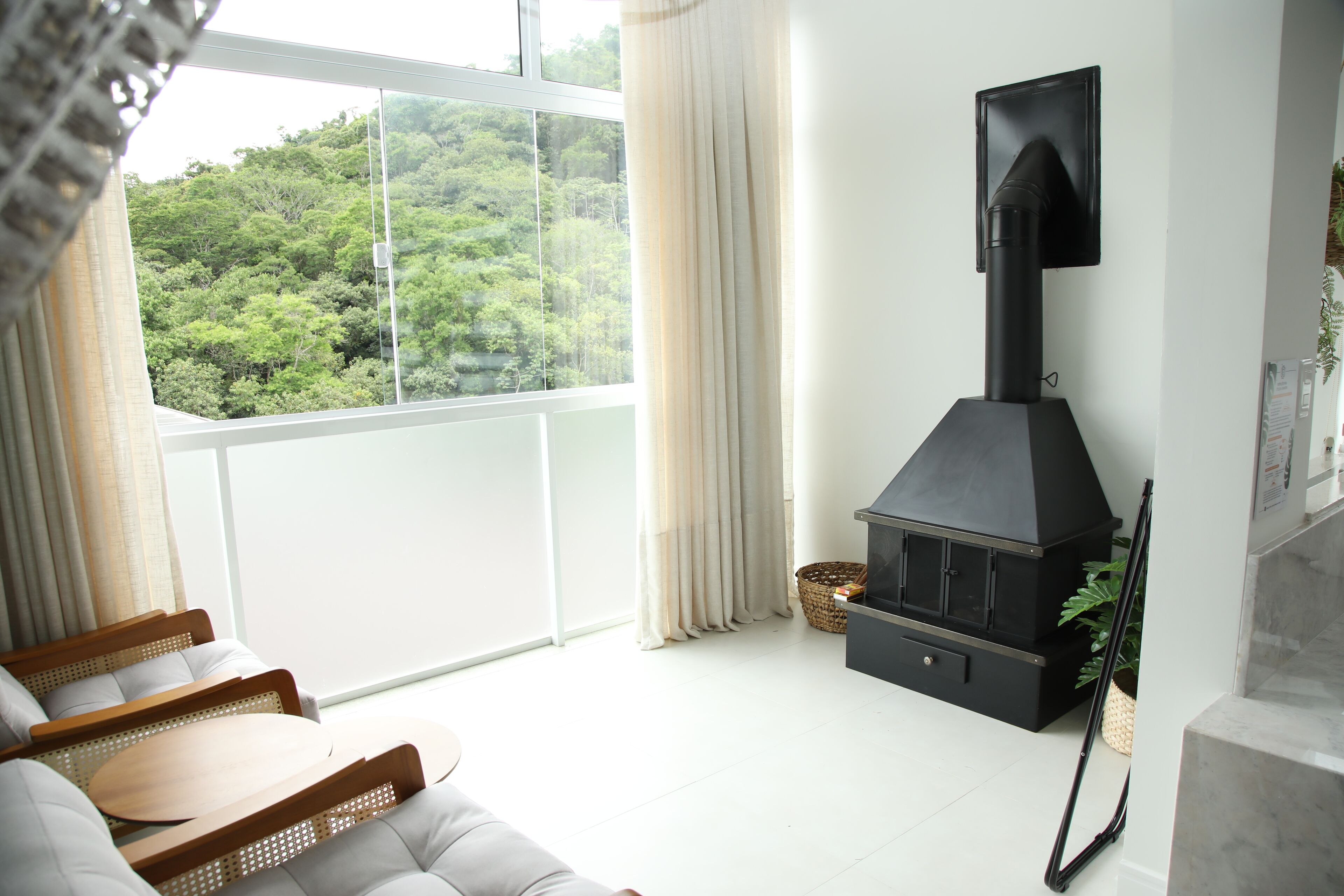 Uma sala de estar moderna com uma janela ampla, uma lareira de madeira preta e vista para uma encosta arborizada.
