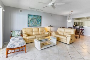 Sala de estar com sofá bege, mesa de centro de vidro e cadeira de madeira. Há um quadro na parede e um ventilador de teto.