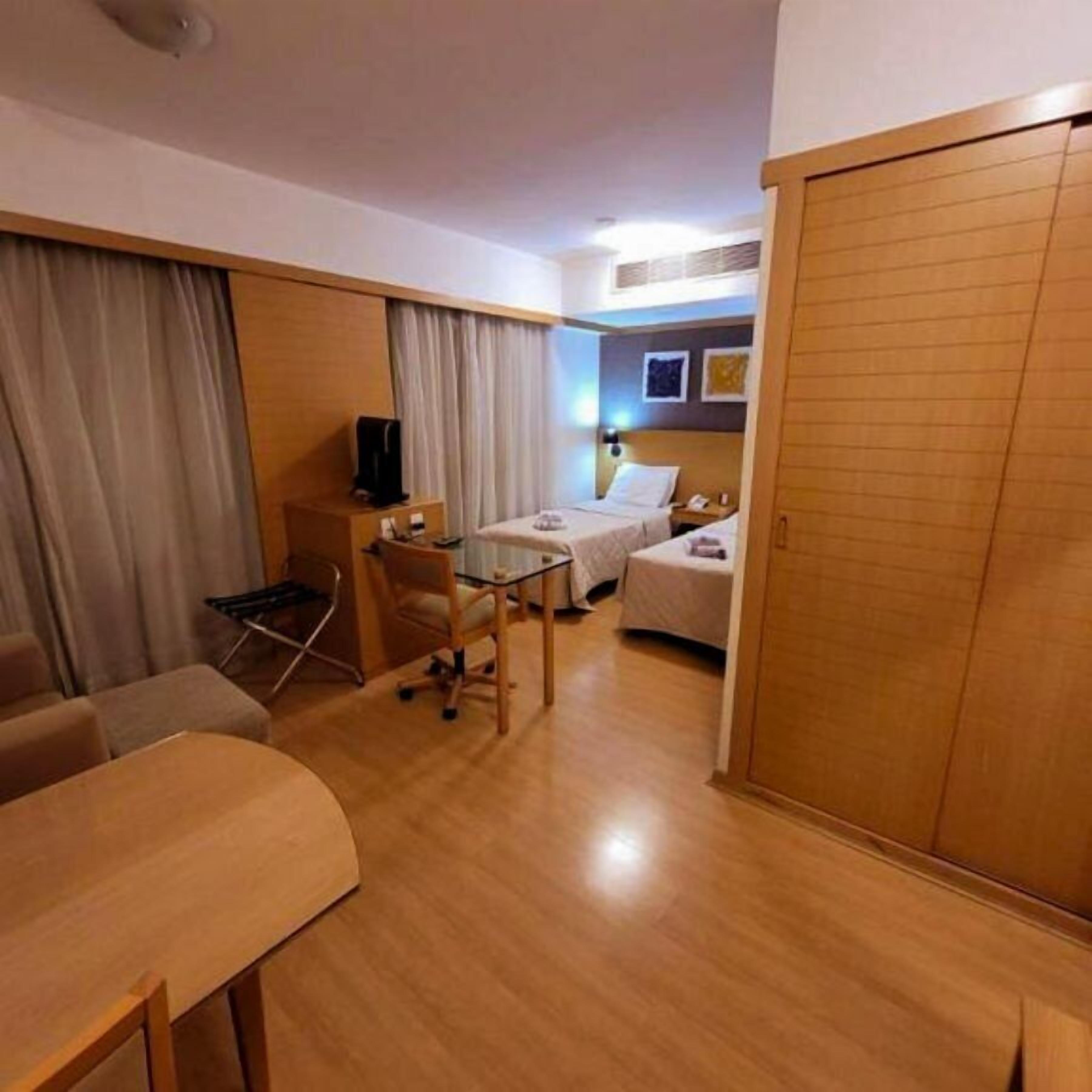 Quarto de hotel pequeno, com uma cama, uma escrivaninha, uma cadeira, uma mesa e um guarda-roupa.