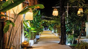 Um caminho noturno ladeado por plantas em vasos e luzes pendentes, conduzindo a uma entrada de edifício.
