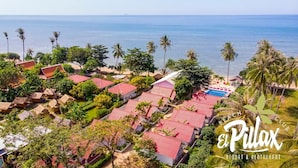 Resort com vários edifícios, alguns com telhados vermelhos, rodeado por vegetação exuberante e palmeiras, com vista para um mar calmo.